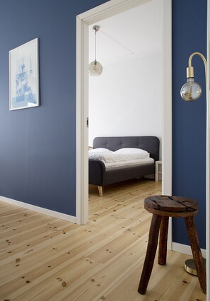 Quarto com cama, um banquinho de madeira, luminária fixada na parede e um quadro emoldurado.
