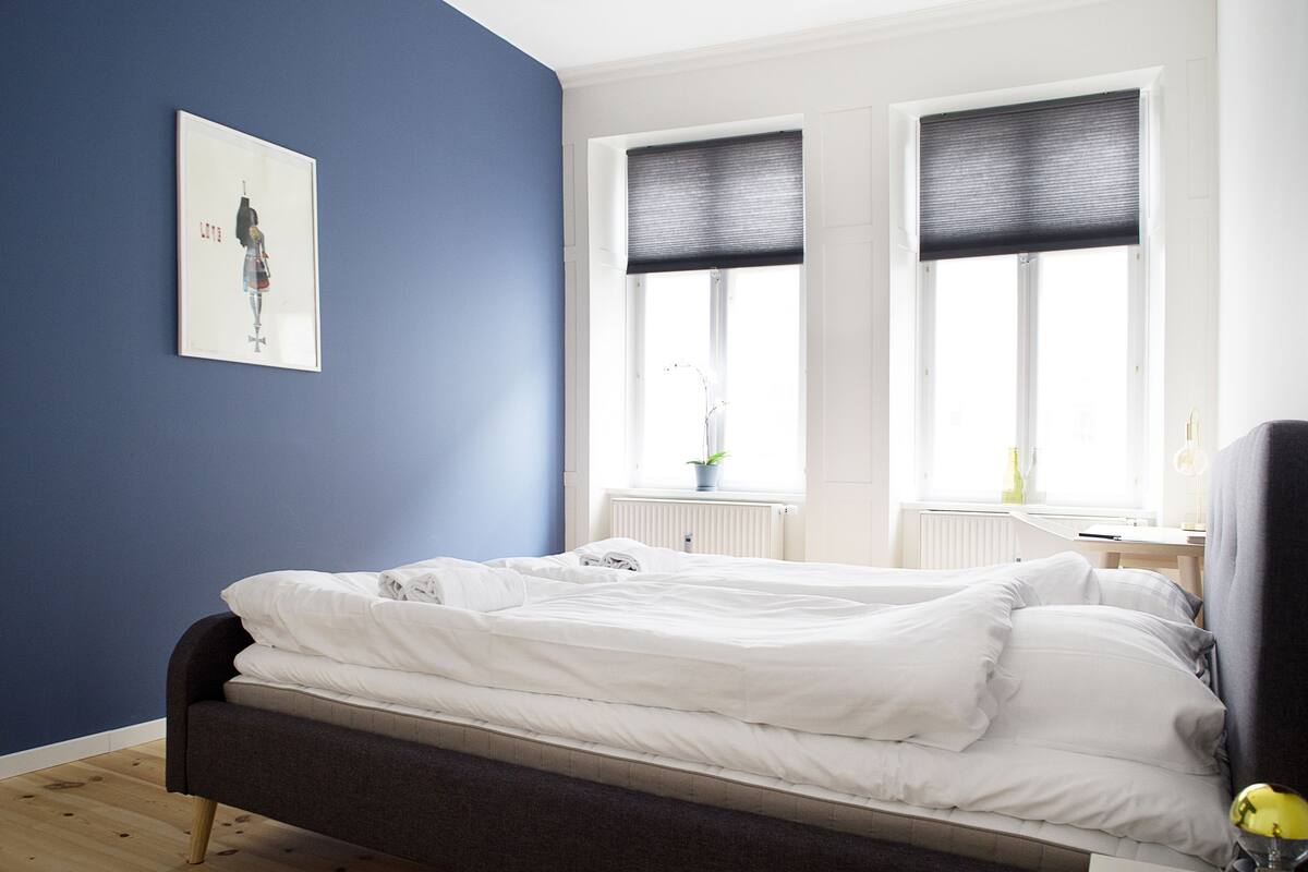 Quarto com uma cama, duas janelas com persianas, um radiador e um quadro na parede.