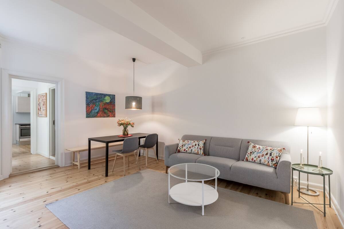 Uma sala de estar moderna com um sofá cinzento, uma mesa de centro redonda, um espaço de jantar com cadeiras, um quadro na parede e um abajur sobre uma mesa lateral.