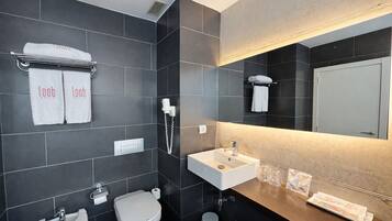Um banheiro moderno com azulejos escuros, vaso sanitário branco, pia, chuveiro manual e porta-toalhas com toalhas.