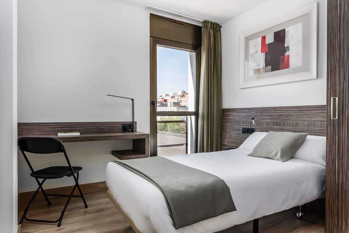 Quarto de hotel com cama, escrivaninha, cadeira, janela com cortinas e vista para a cidade.
