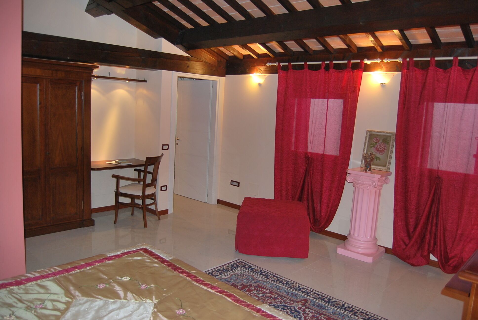 Um quarto com escrivaninha de madeira, uma cadeira, uma cama com colcha vermelha, mesa de cabeceira rosa com uma fotografia e cortinas vermelhas.