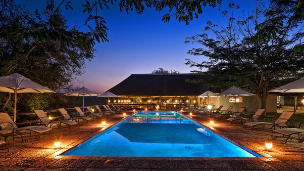 Área na piscina com espreguiçadeiras e guarda-sóis, cercada por árvores e um edifício ao fundo.