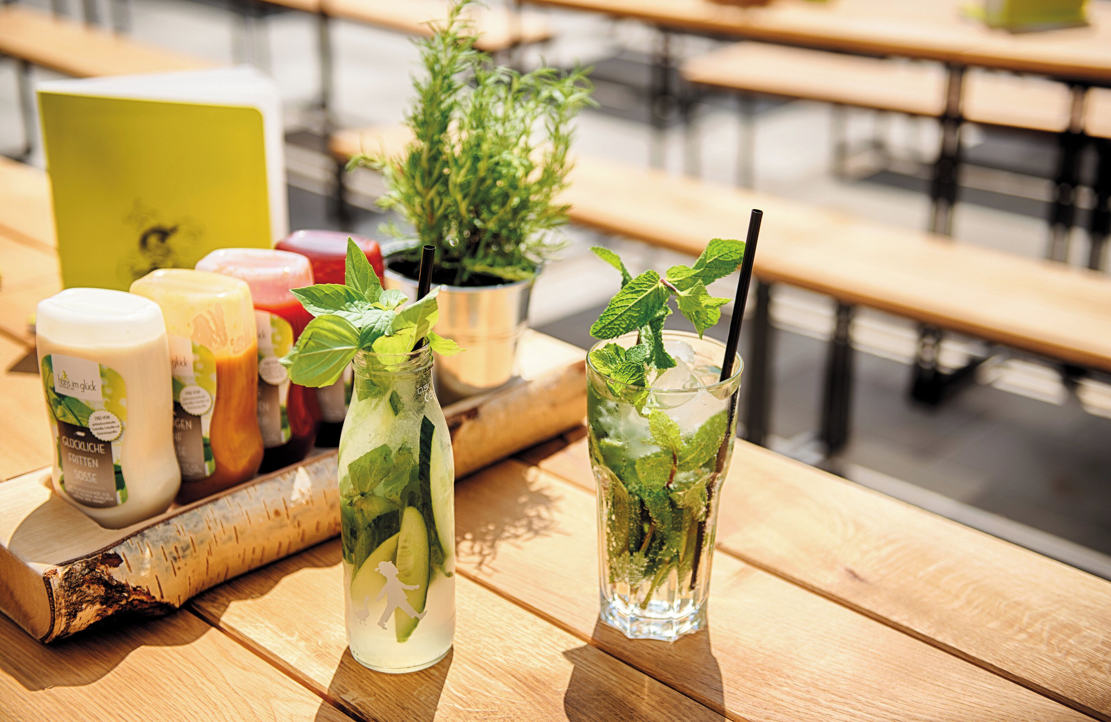 Uma mesa de madeira com três bebidas, um prato de acompanhamentos e uma planta em vaso.