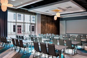 Sala de conferência com mesas, cadeiras, janelas amplas e tela de projeção.