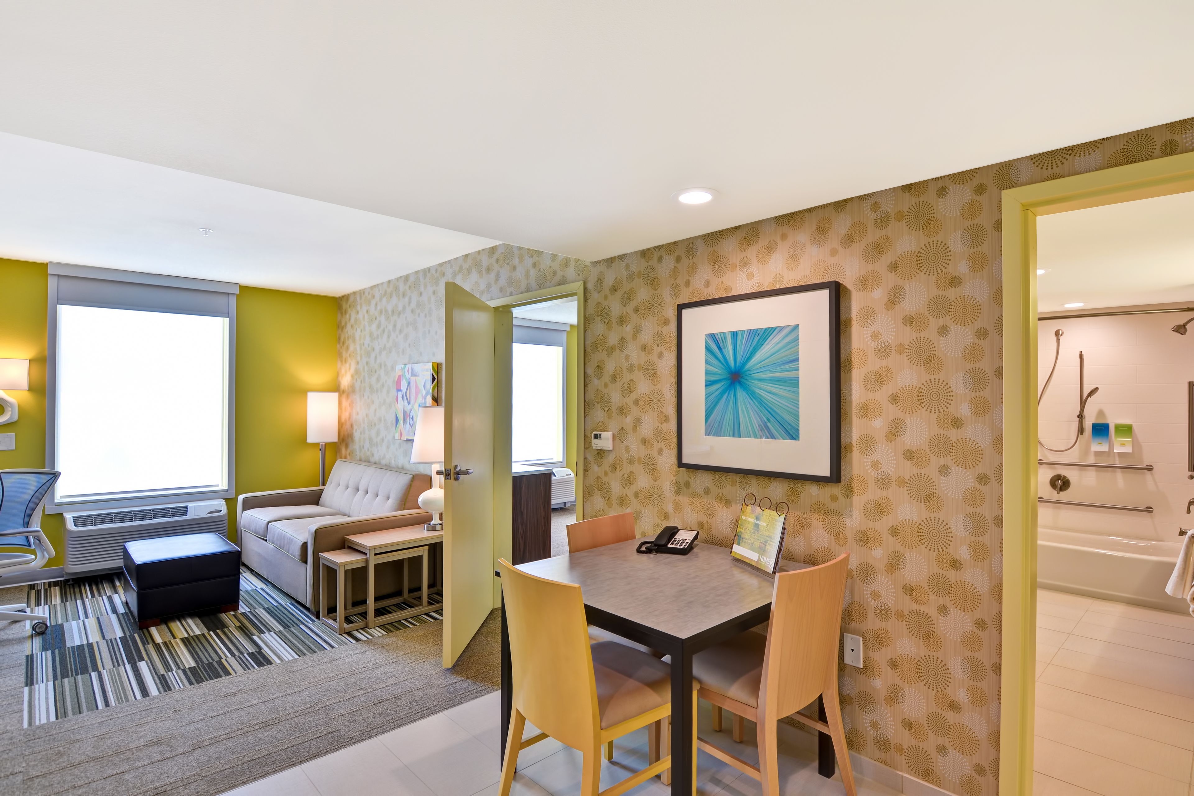 Quarto de hotel com cama, escrivaninha, cadeira, abajur, quadro e banheiro.