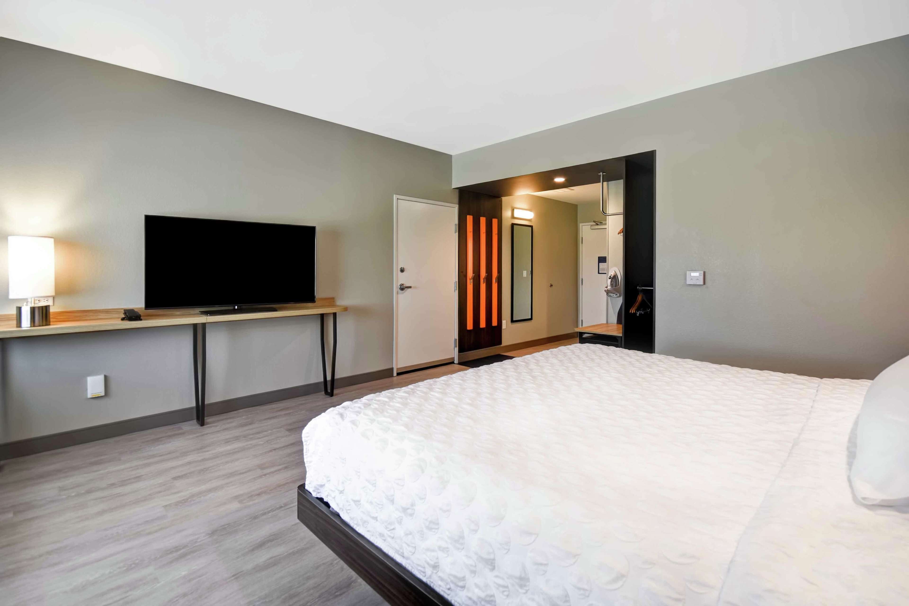 Quarto de hotel moderno com uma cama grande, televisão de tela plana, mesa de cabeceira e espelho.