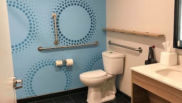 Casa de banho com parede azul com padrão circular, sanita branca, barra de apoio, lavatório com espelho e um contentor de lixo vermelho.
