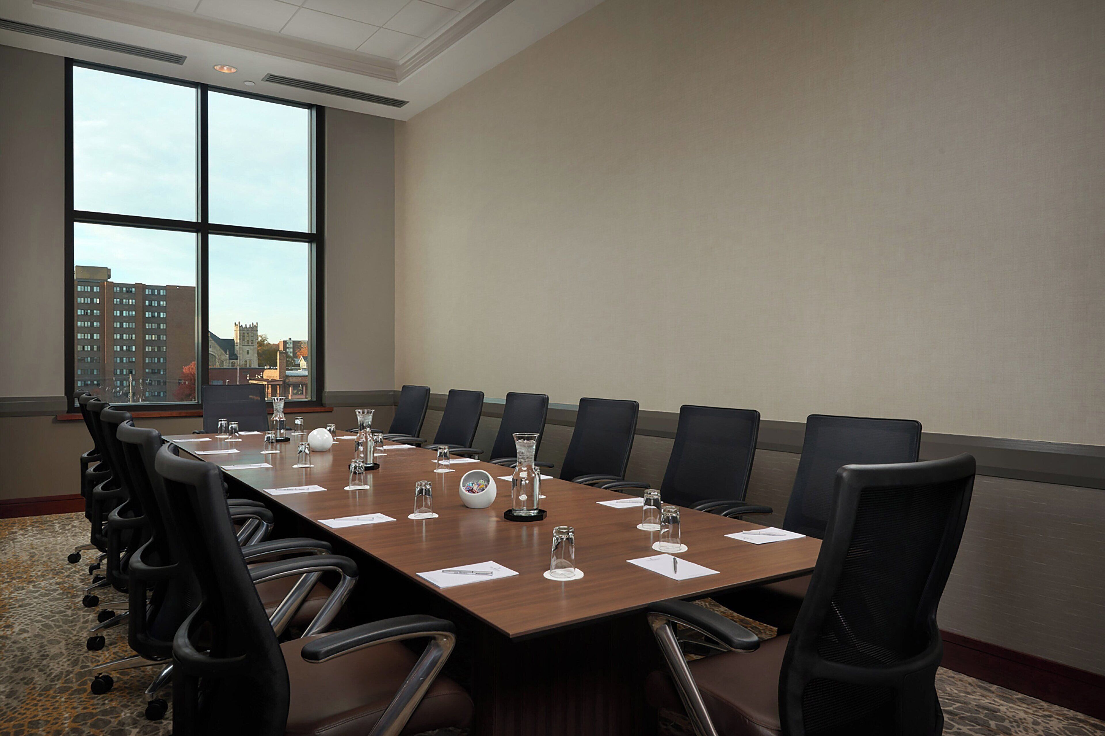 Sala de conferências com uma longa mesa, cadeiras pretas e vista para a cidade através de amplas janelas.