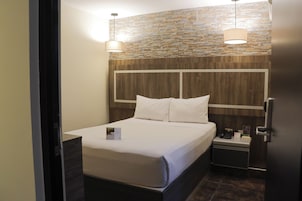 Quarto de hotel moderno com uma cama bem arrumada, mesa de cabeceira com telefone e luminária, e parede texturizada em pedra atrás da cama.