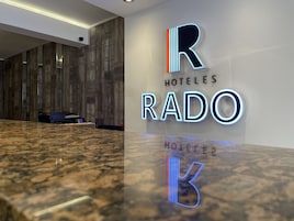 Uma área de recepção de hotel com balcão de mármore, parede de madeira e sinalização iluminada para 'HOTELES RADO'.