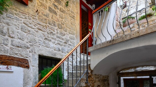 Uma escadaria de pedra leva a uma porta de madeira com uma placa de 'Welcome', corrimão de metal e uma pequena janela com vegetação.