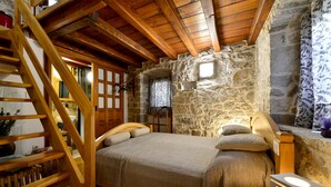 Quarto rústico com escada de madeira, paredes de pedra, cama com colcha estampada e uma pequena janela.