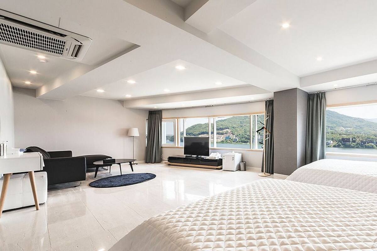 Quarto de hotel moderno com duas camas, uma escrivaninha, uma cadeira, televisão e janelas amplas com vista para o verde.