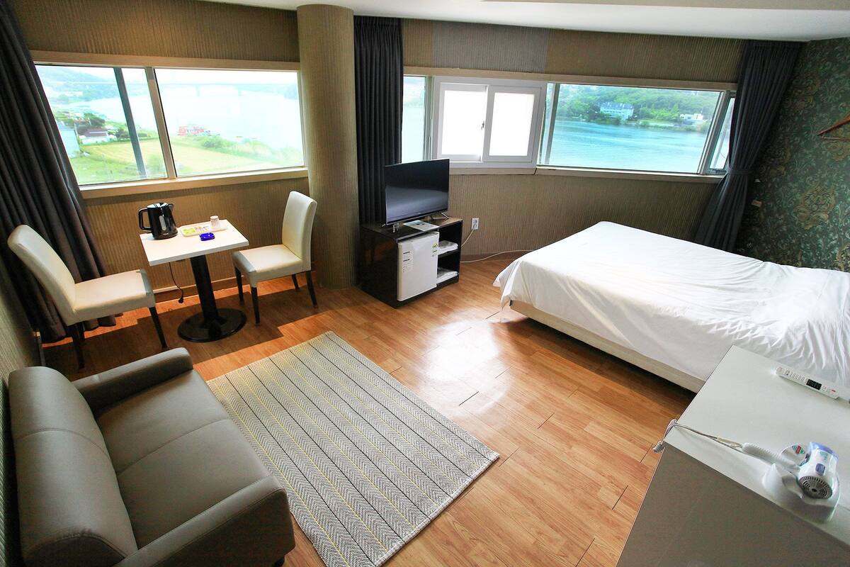 Quarto de hotel com uma cama, escrivaninha com televisão, sofá e vista para um corpo de água.