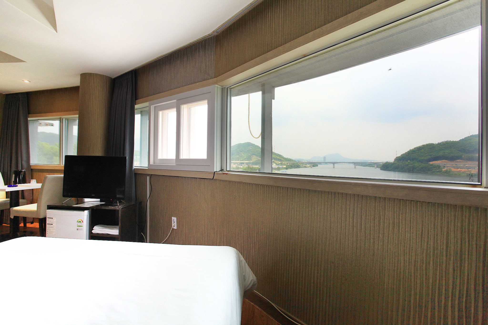 Quarto de hotel com cama, escrivaninha, televisão e janela com vista para uma ponte e colinas.