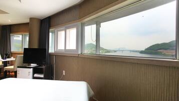 Quarto de hotel com cama, escrivaninha, televisão e janela com vista para uma ponte e colinas.