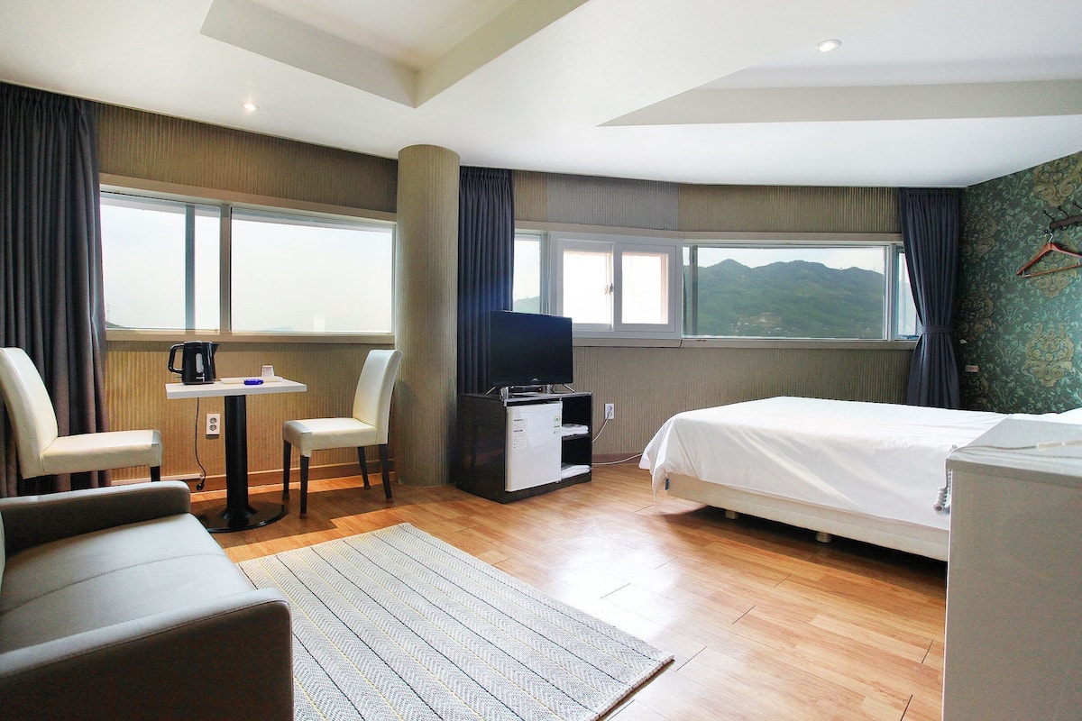 Quarto de hotel com cama, escrivaninha com cadeira, televisão e vista para montanhas pelas janelas.