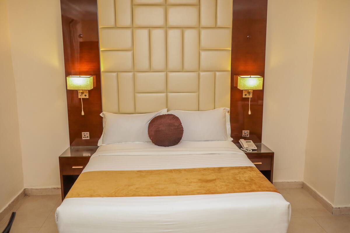 Quarto de hotel com uma cama grande, duas mesas de cabeceira, cabeceira com painel de luz e um telefone em uma das mesas.