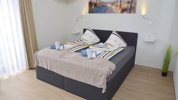 Quarto moderno com cama escura, almofadas listradas, manta bege e arte na parede com tema praiano.