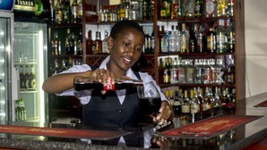 Um barman servindo uma bebida em um bar, com prateleiras cheias de garrafas de bebidas alcoólicas ao fundo.