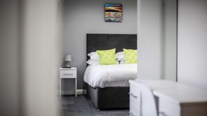 Quarto moderno com cabeceira cinza, cama com almofadas estampadas em branco e verde, mesa de cabeceira com abajur e um quadro na parede.