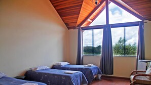 Quarto com três camas, soalho de madeira, uma janela grande com cortinas e uma cadeira de vime.
