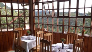 Uma sala de jantar com mesas e cadeiras de madeira, grandes janelas com vista para uma paisagem arborizada.