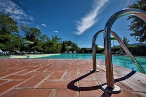 Uma piscina com deck ladrilhado, guarda-corpos de metal na beira e espreguiçadeiras sob guarda-sóis.