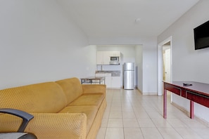 Espaço compacto com sofá, uma mesinha, cozinha equipada com micro-ondas e frigorífico, e televisão na parede.