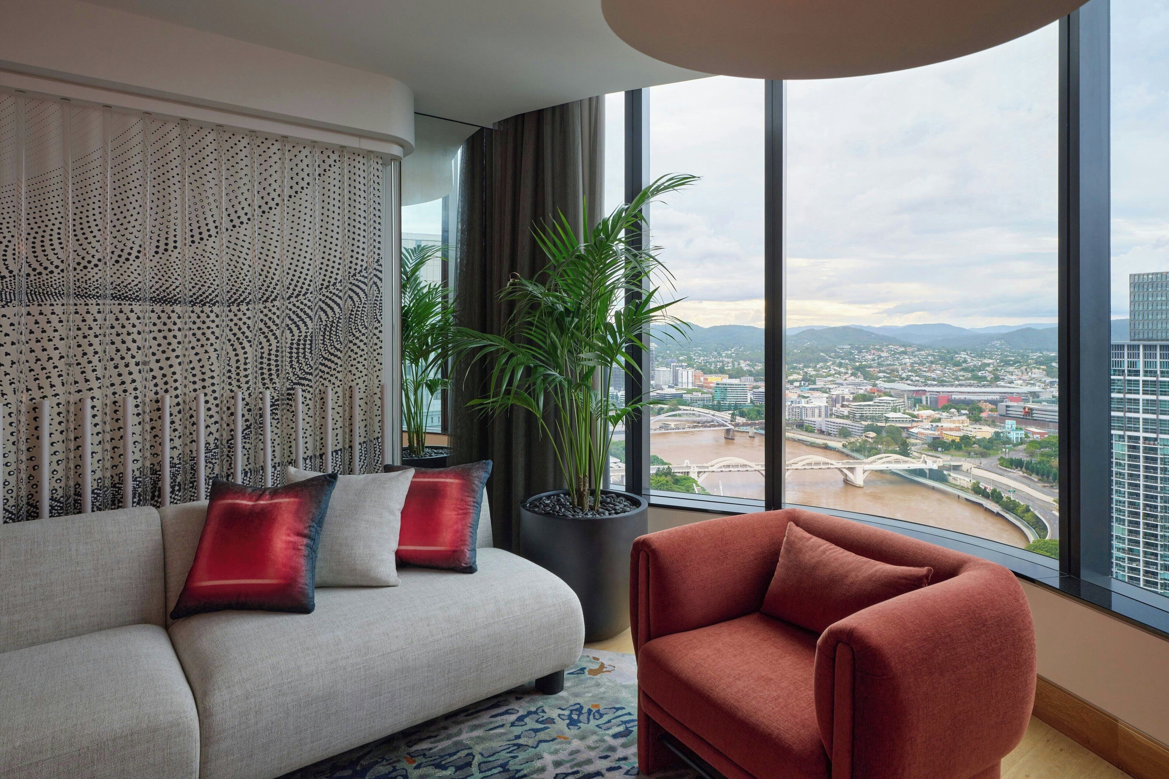 Uma sala de estar moderna com um sofá, uma poltrona vermelha e uma grande planta em vaso. Um painel decorativo e uma vista da cidade e do rio são visíveis através de grandes janelas.