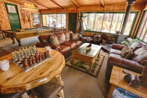 Interior acolhedor de uma cabana com mesa de bilhar, sofás de couro, um tabuleiro de xadrez e lareira.