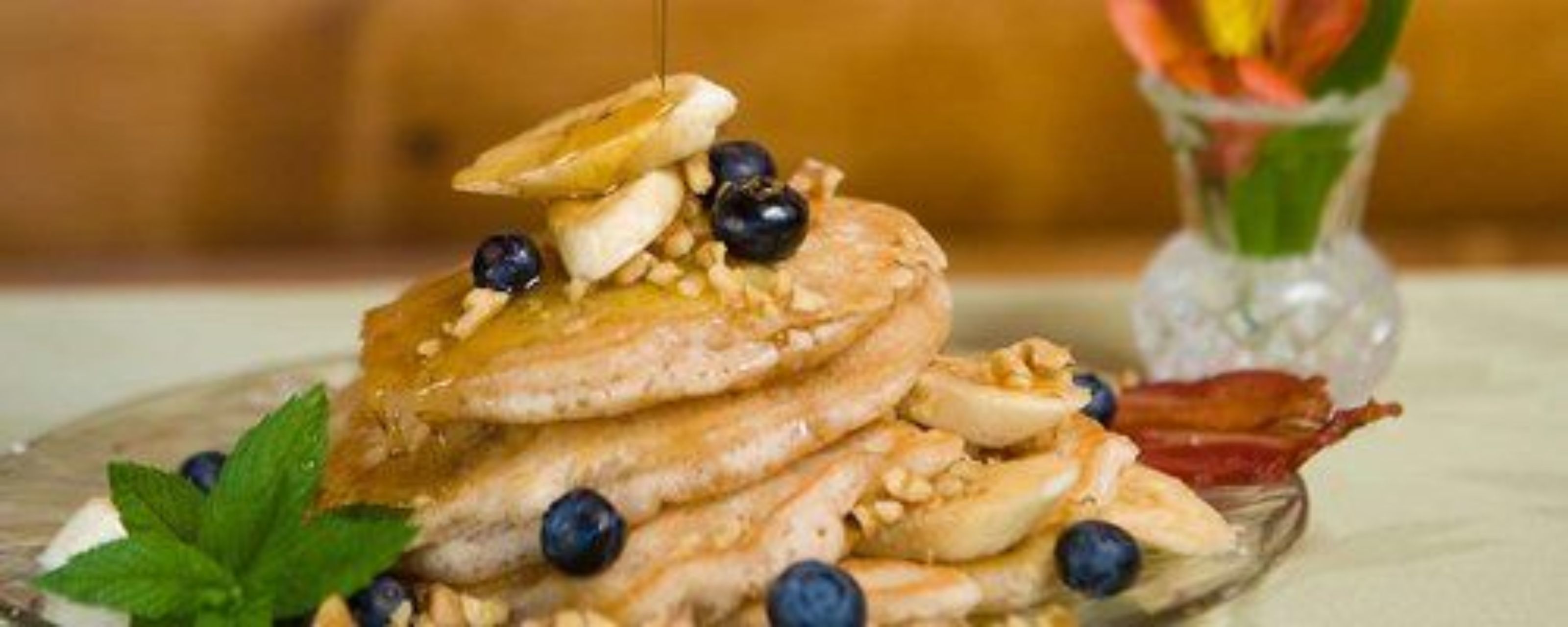 Uma pilha de panquecas com amoras e nozes, calda sendo despejada por cima, e um copo de suco ao fundo.