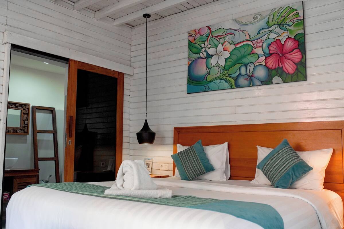 Quarto de hotel com cabeceira de madeira, cama com roupa de cama branca e verde-água, pintura de flores tropicais e escrivaninha de madeira com cadeira.