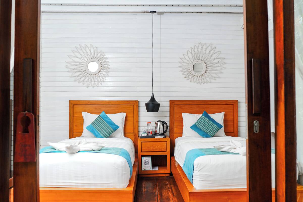 Quarto duplo com duas camas, cabeceiras de madeira, roupa de cama branca e mesa de cabeceira com chaleira.