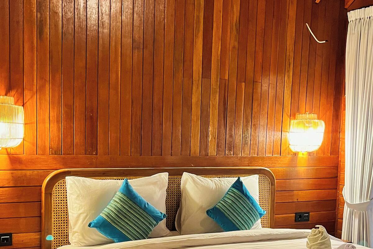 Quarto com painéis de madeira, uma cama, almofadas, mesa de cabeceira e luminárias fixadas na parede.