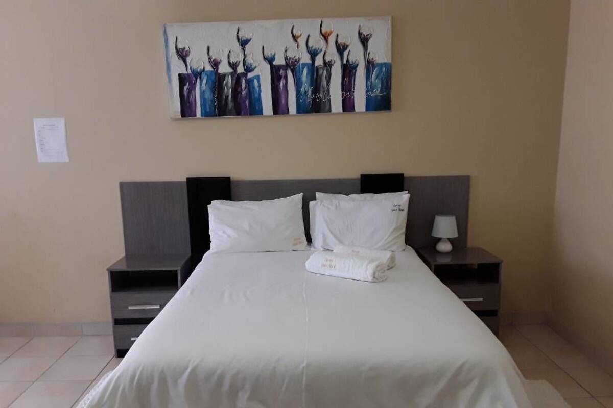 Quarto de hotel com uma cama grande, duas mesas de cabeceira, um criado-mudo, um quadro na parede e uma toalha branca na cama.
