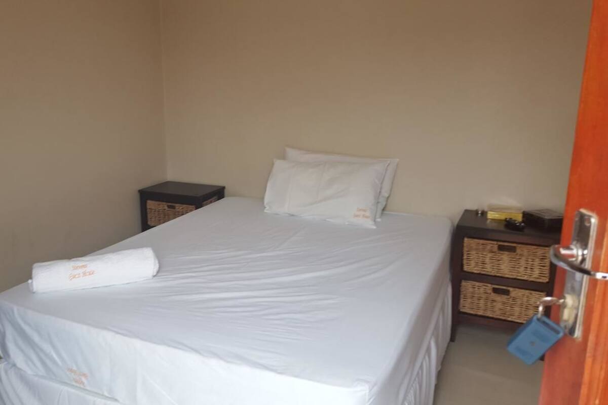 Cama bem arrumada com lençóis brancos e uma toalha dobrada, mesa de cabeceira com abajur e um criado-mudo com um cesto.