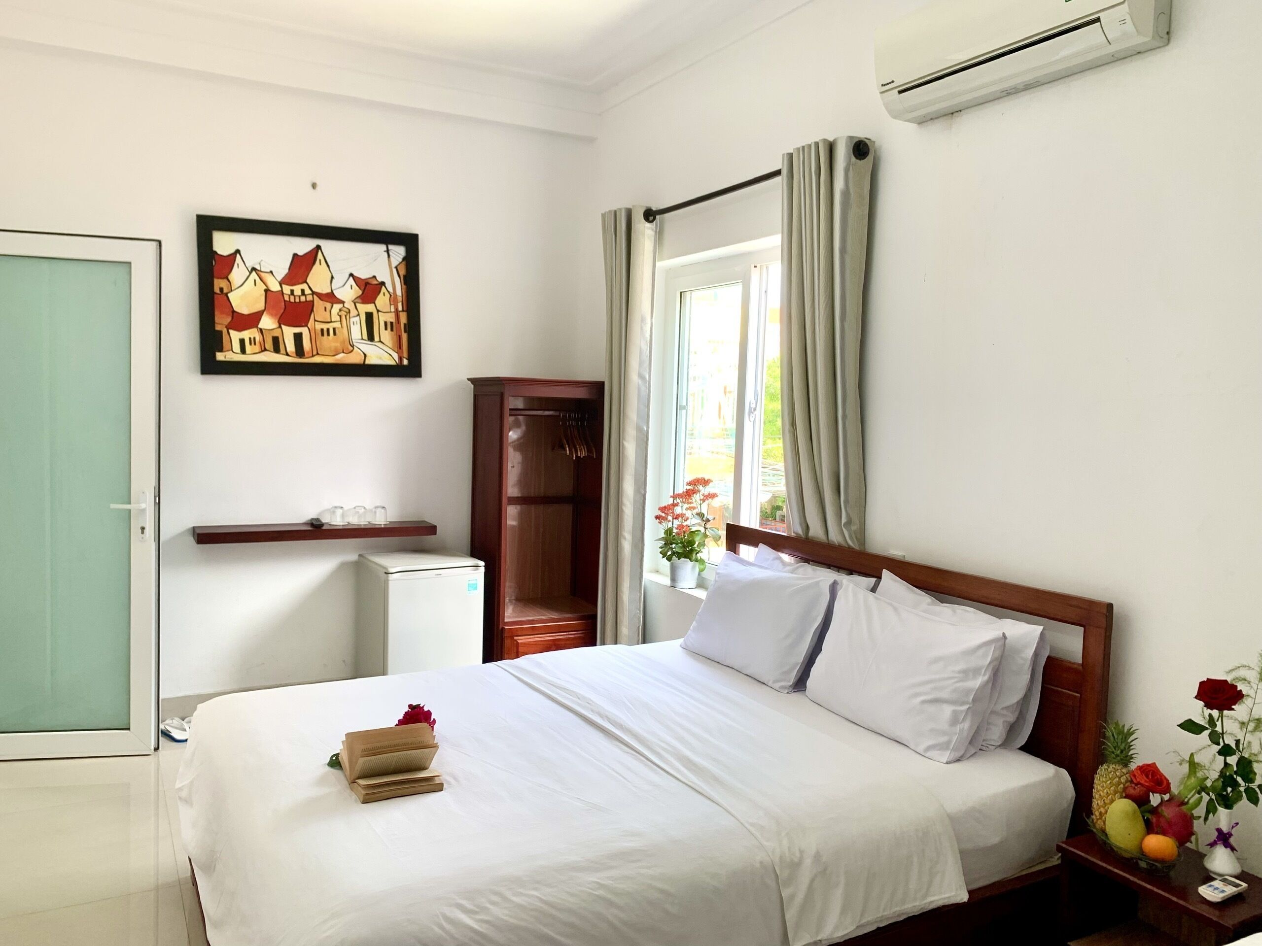 Quarto de hotel com cama, guarda-roupa, mini-frigorífico, janela com cortinas, um quadro na parede e uma planta no peitoril.