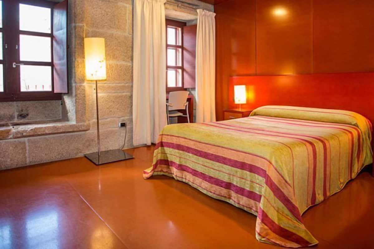 Um quarto com uma cama, uma mesa de cabeceira, uma luminária e uma janela com cortinas.