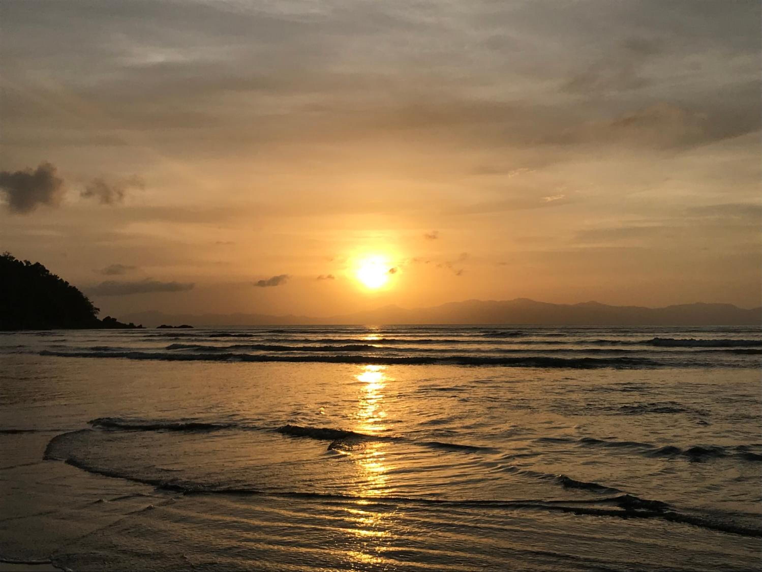 Pôr do sol sobre uma praia com ondas e uma cadeia montanhosa ao longe.