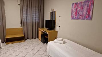 Quarto de hotel com cama, escrivaninha com televisão, cadeira, banco e janela com cortinas.