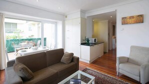 Uma sala de estar moderna com um sofá marrom, uma mesa de centro de vidro e uma poltrona bege. Uma cozinha é visível ao fundo, com armários brancos e um micro-ondas.