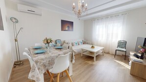 Uma sala de jantar com mesa posta para quatro pessoas, um sofá, uma TV e um abajur.