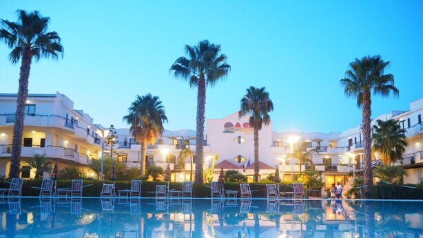 Área de piscina com espreguiçadeiras e palmeiras, cercada por edifícios altos com varandas e uma placa de hotel visível.