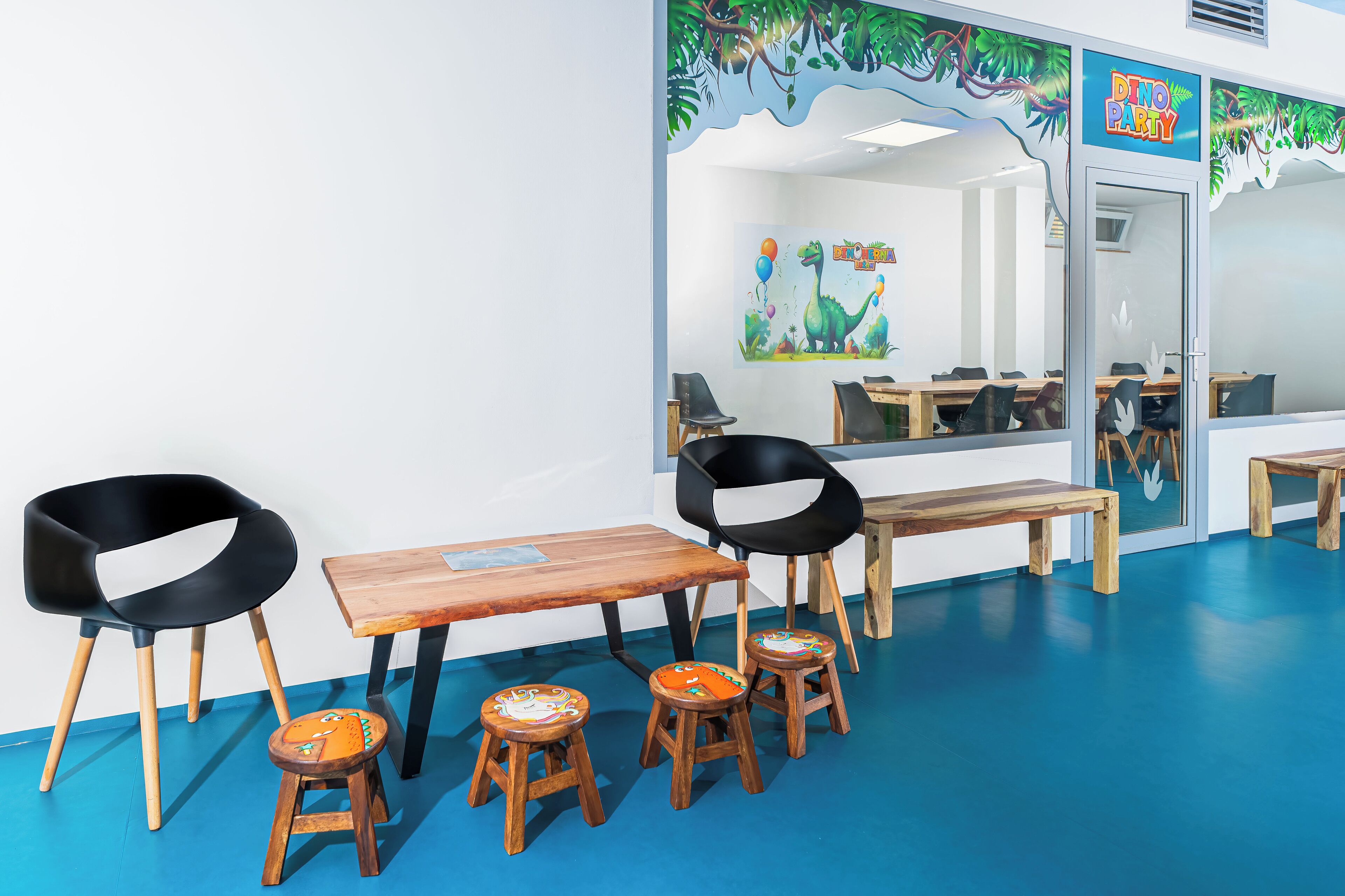 Um quarto com piso azul, mesas de madeira, cadeiras pretas e de madeira, e um mural com uma festa de dinossauros.