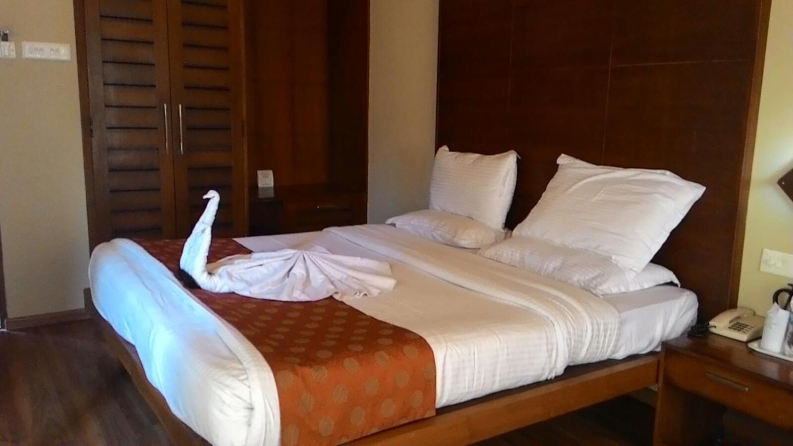 Quarto de hotel com cama de madeira, telefone na mesa de cabeceira e porta com puxador.