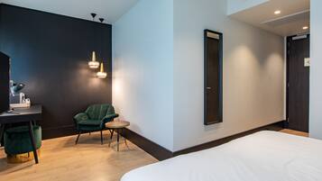 Quarto de hotel moderno com uma cama grande, uma escrivaninha com cadeira, uma mesa redonda pequena e uma poltrona verde.