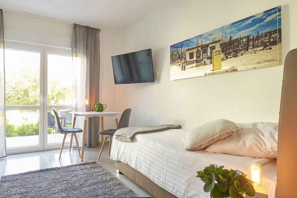 Quarto de hotel com cama, escrivaninha, cadeira, televisão e uma grande janela.