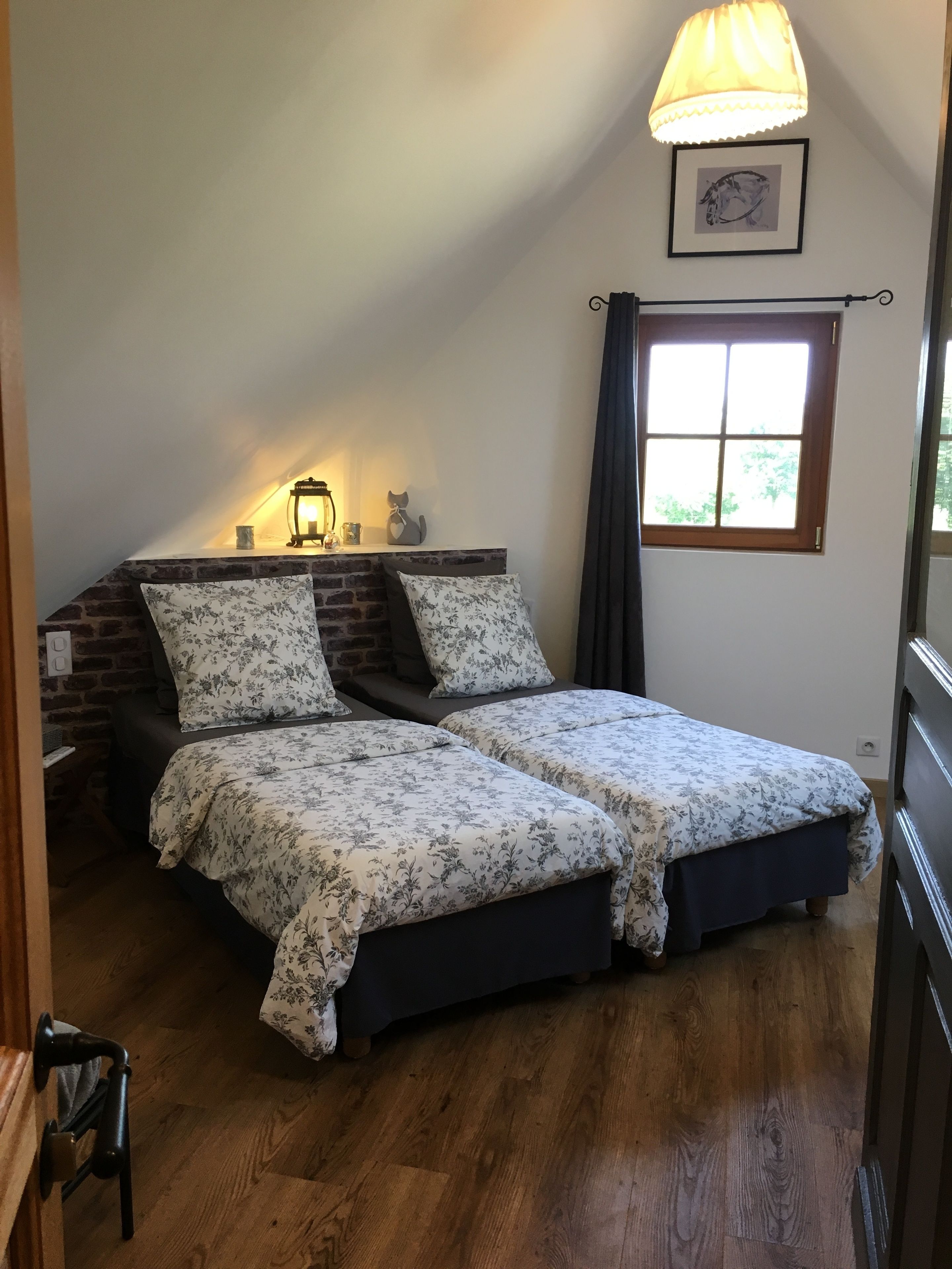 Um quarto com parede de tijolos, janela com cortinas, cama com roupa de cama florida e luminária fixada na parede.
